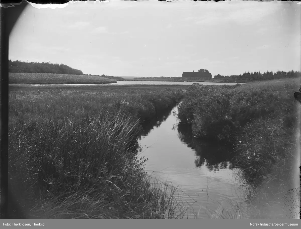 Hus og driftsbygg, elv og liten bekk i forgrunnen. Sannsynligvis fra ...