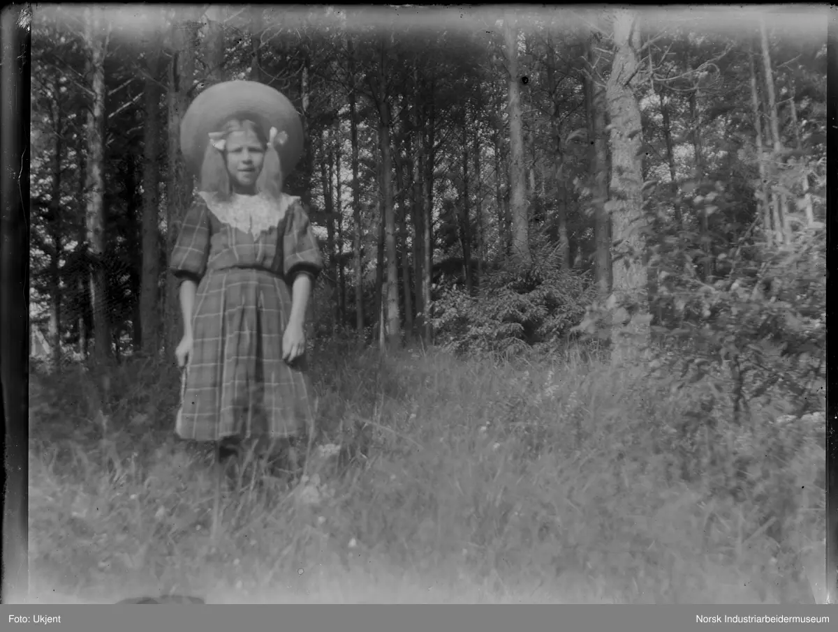 Jente stående i tett granskog. Jenta er kledd i rutet kjole med ...