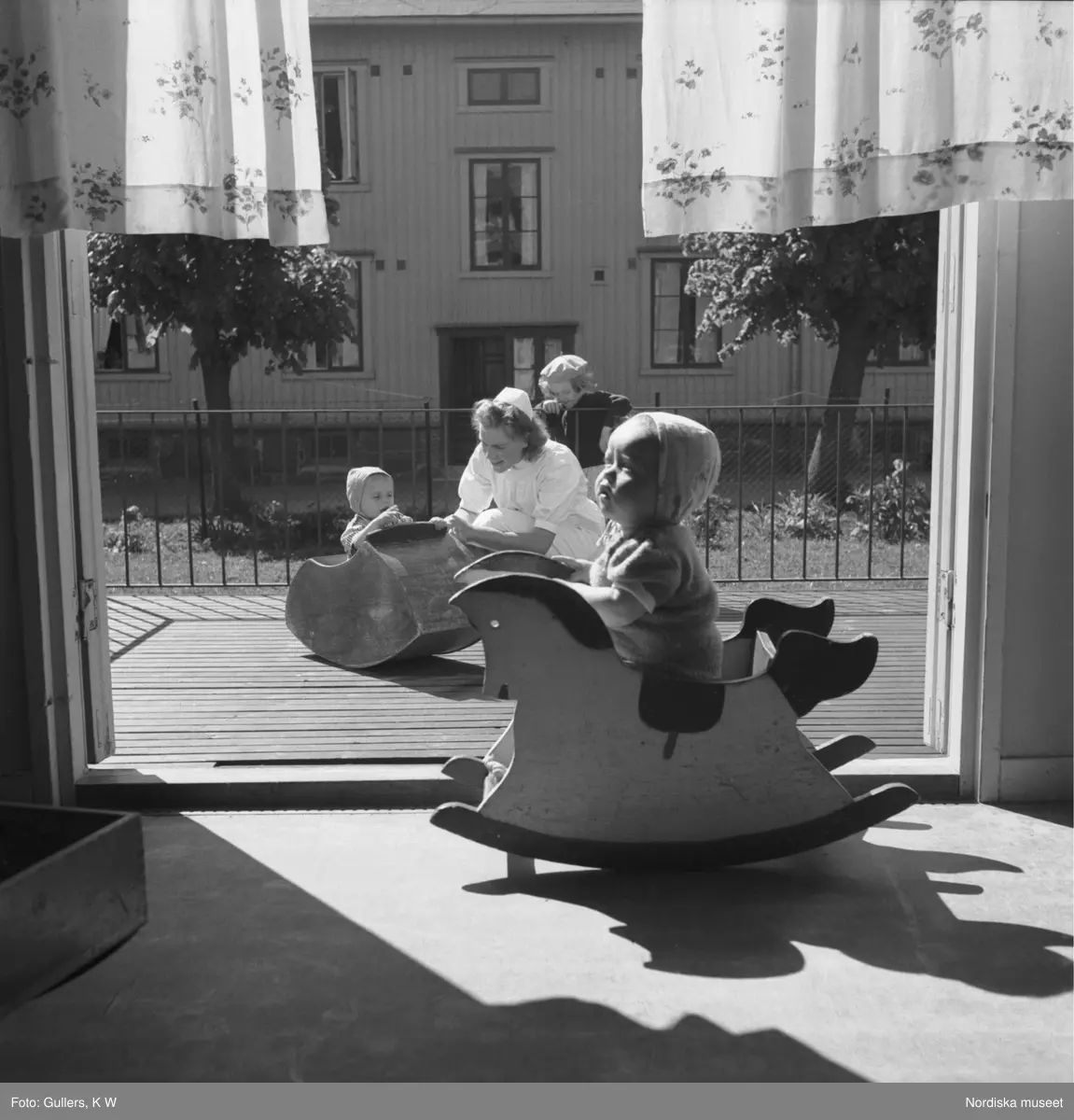 Tre små barn och en vitklädd kvinna i en liten vit hatt. Bilden är från ...