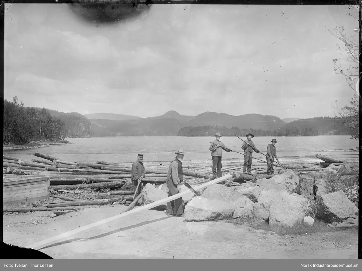 Fem tømmerfløtere med fløteredskap og sag ved elvebredd. Tømmer ligger ...